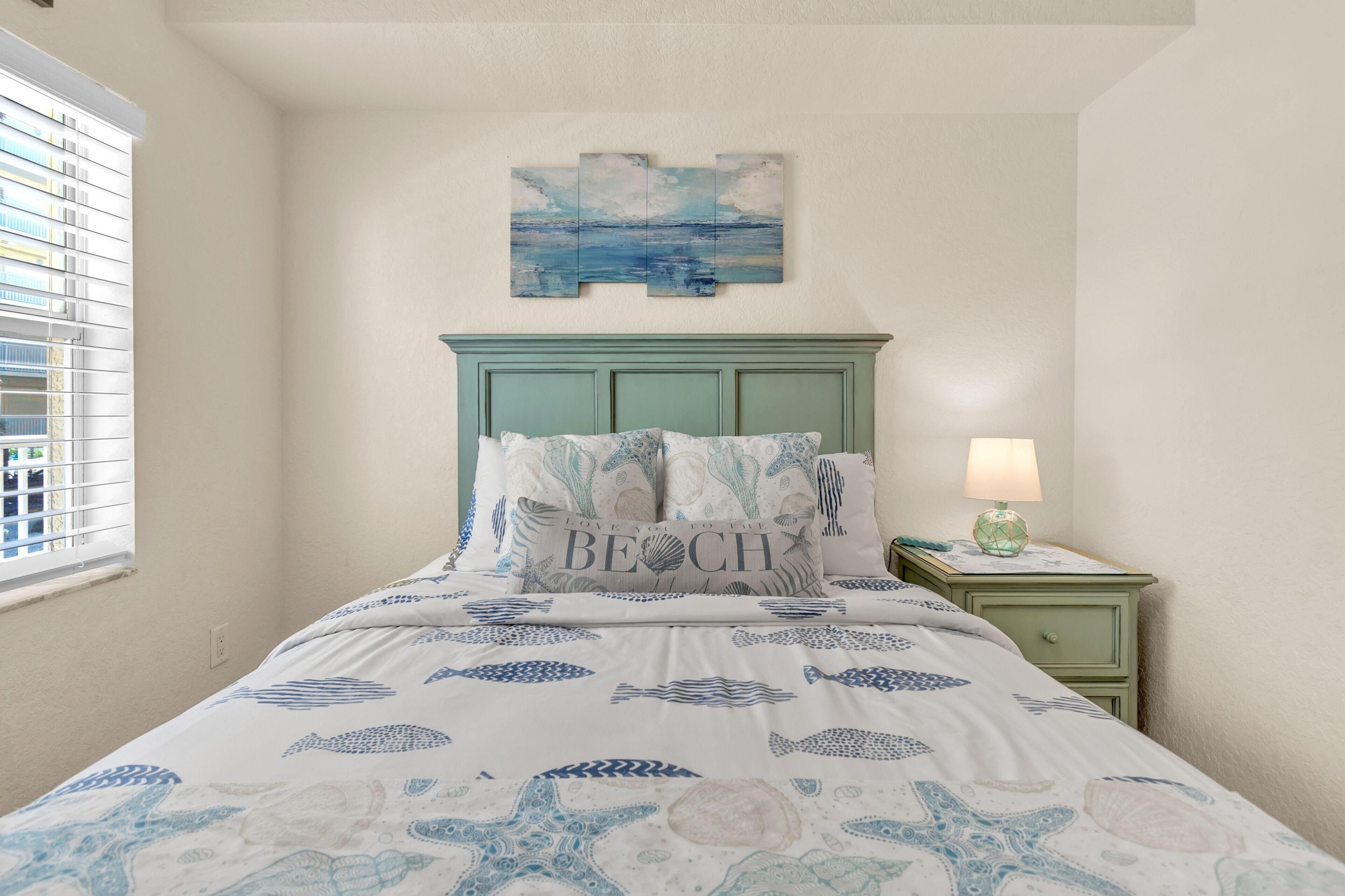 Quarto com cama temática náutica, mesa de cabeceira com abajur e janela com persianas.