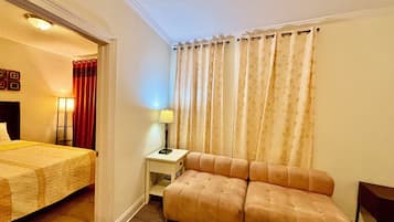 Quarto de hotel com cama, sofá, mesa de cabeceira com abajur e janela com cortinas.