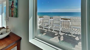 Quarto com uma grande janela com vista para uma praia, com cadeiras brancas e um deck de madeira.
