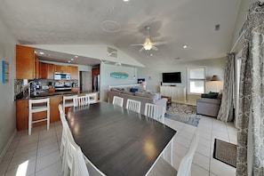 Cozinha moderna com ilha central, sala de jantar com mesa escura e cadeiras brancas, sala de estar com sofá e TV de tela plana, e ventilador de teto.