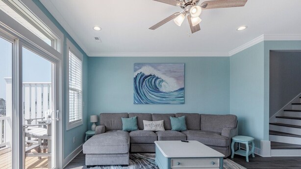 Sala de estar com sofá cinza, mesa de centro branca e tapete azul. Há um quadro de uma onda na parede e um ventilador de teto.
