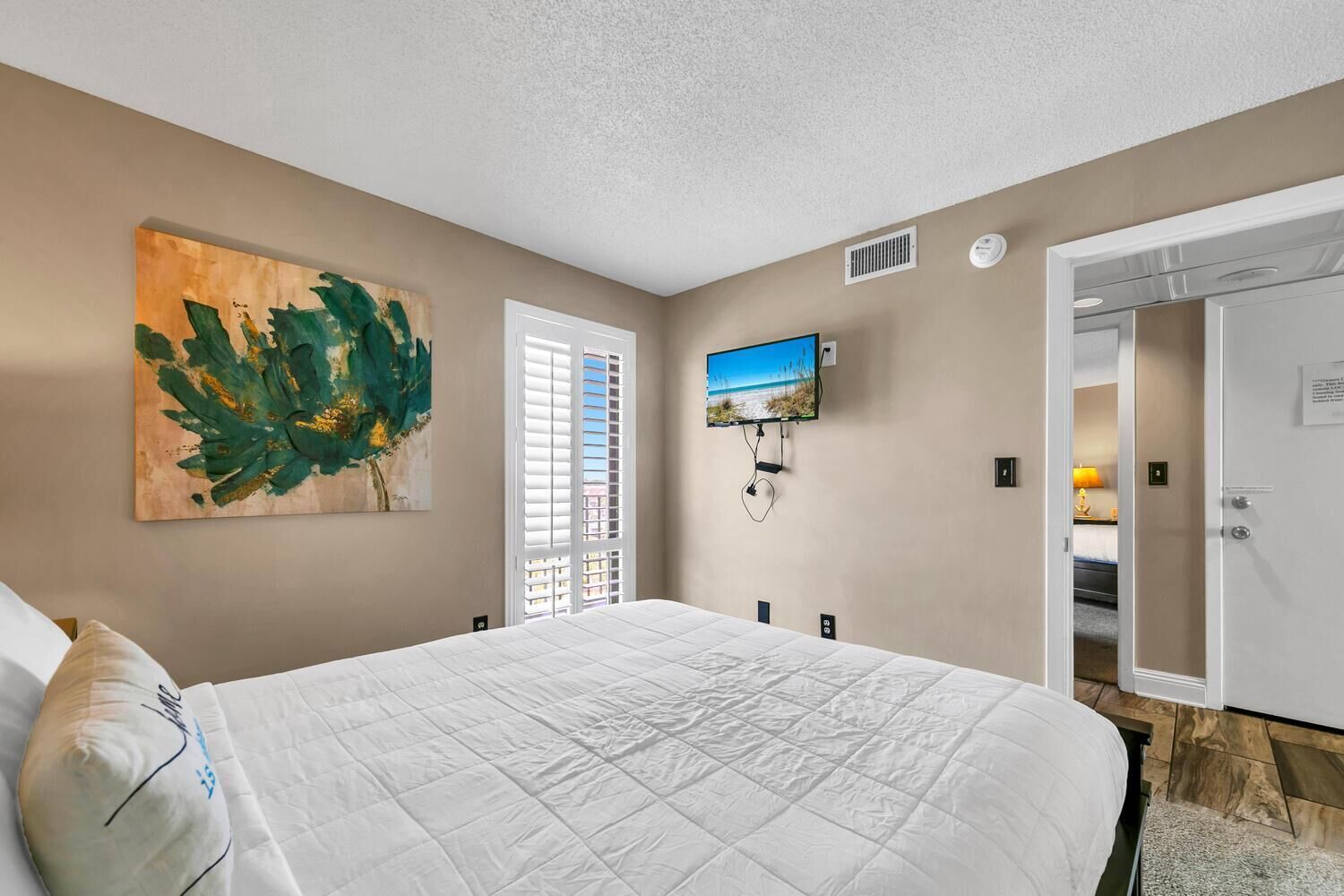Quarto com uma cama grande, um quadro na parede, uma televisão montada acima e uma porta que dá para outro cômodo.