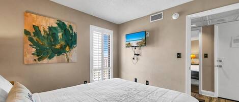 Quarto com uma cama grande, um quadro na parede, uma televisão montada acima e uma porta que dá para outro cômodo.