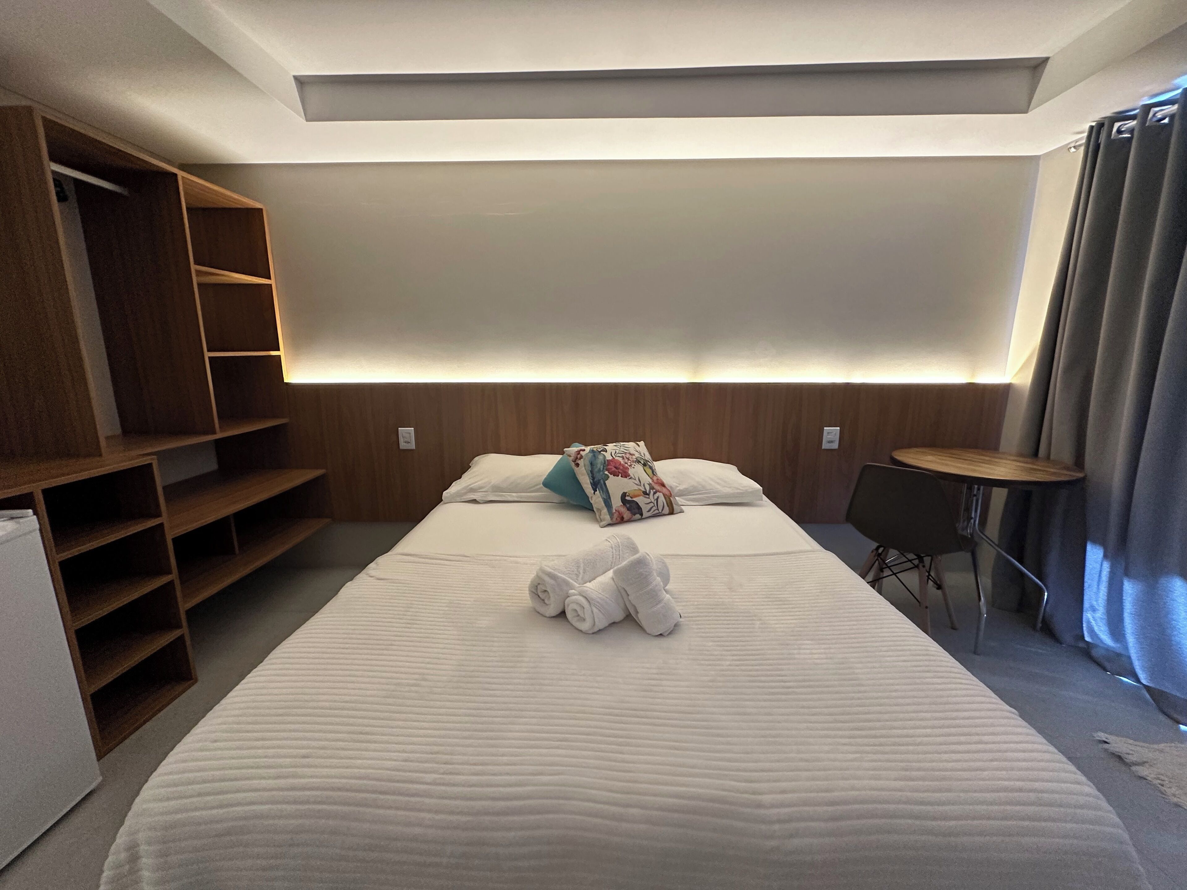 Quarto de hotel moderno com uma cama grande, um guarda-roupa de madeira, uma pequena mesa e uma escrivaninha.