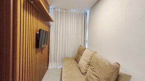 Quarto com parede de painel de madeira, televisão montada, sofá bege e janela com cortinas leves.