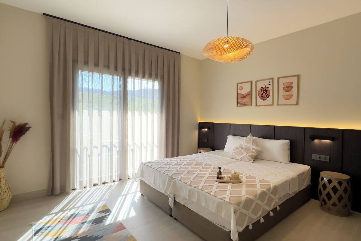 Quarto moderno com cama grande, abajur e vista para o exterior através de cortinas leves.