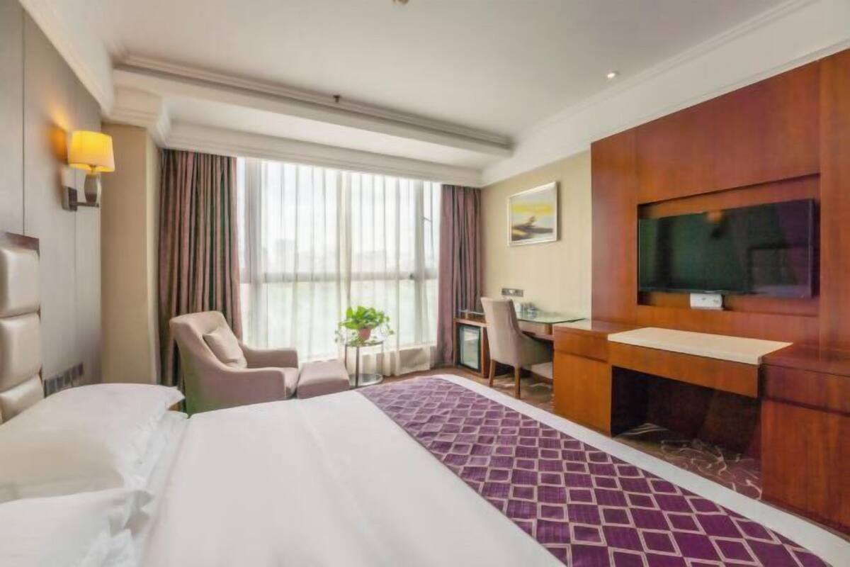 Quarto de hotel com cama, escrivaninha, cadeira, televisão e janela com cortinas.