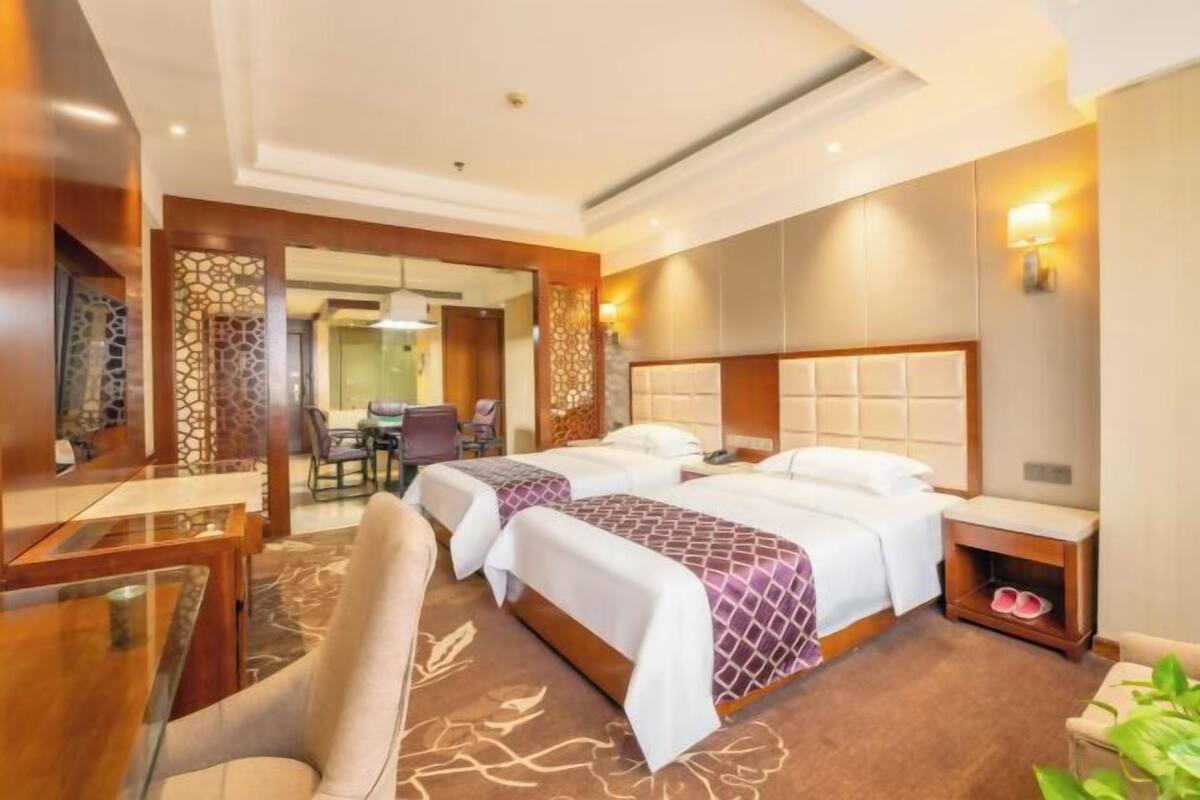 Quarto de hotel com duas camas, uma escrivaninha, uma cadeira, uma mesa pequena e uma mesinha de cabeceira.