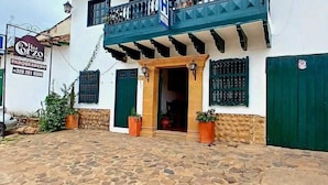 Um edifício de dois andares com varanda, um letreiro do 'El Corzo Hotel' e uma rua de paralelepípedos na frente.