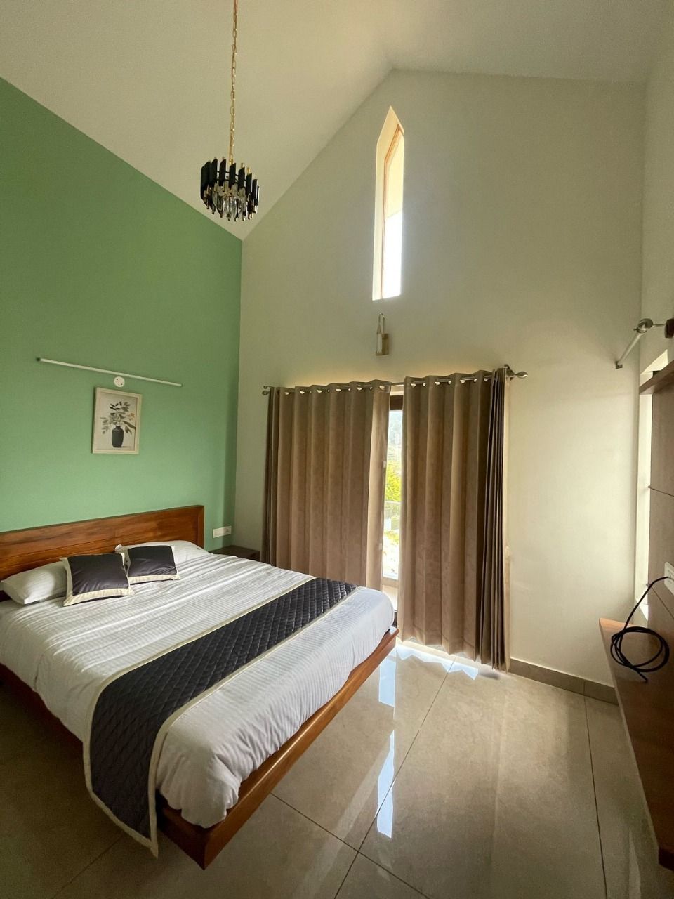 Quarto com cama, luminária pendente, janela com cortinas e um pequeno quadro na parede.