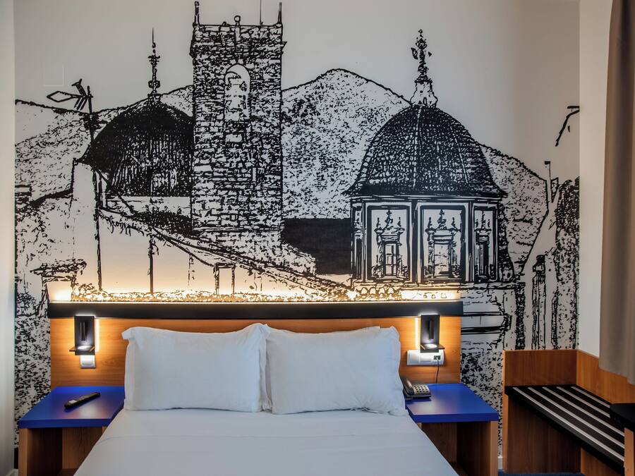 Quarto de hotel com cama, mesas de cabeceira, luminária fixada na parede e um mural decorativo.