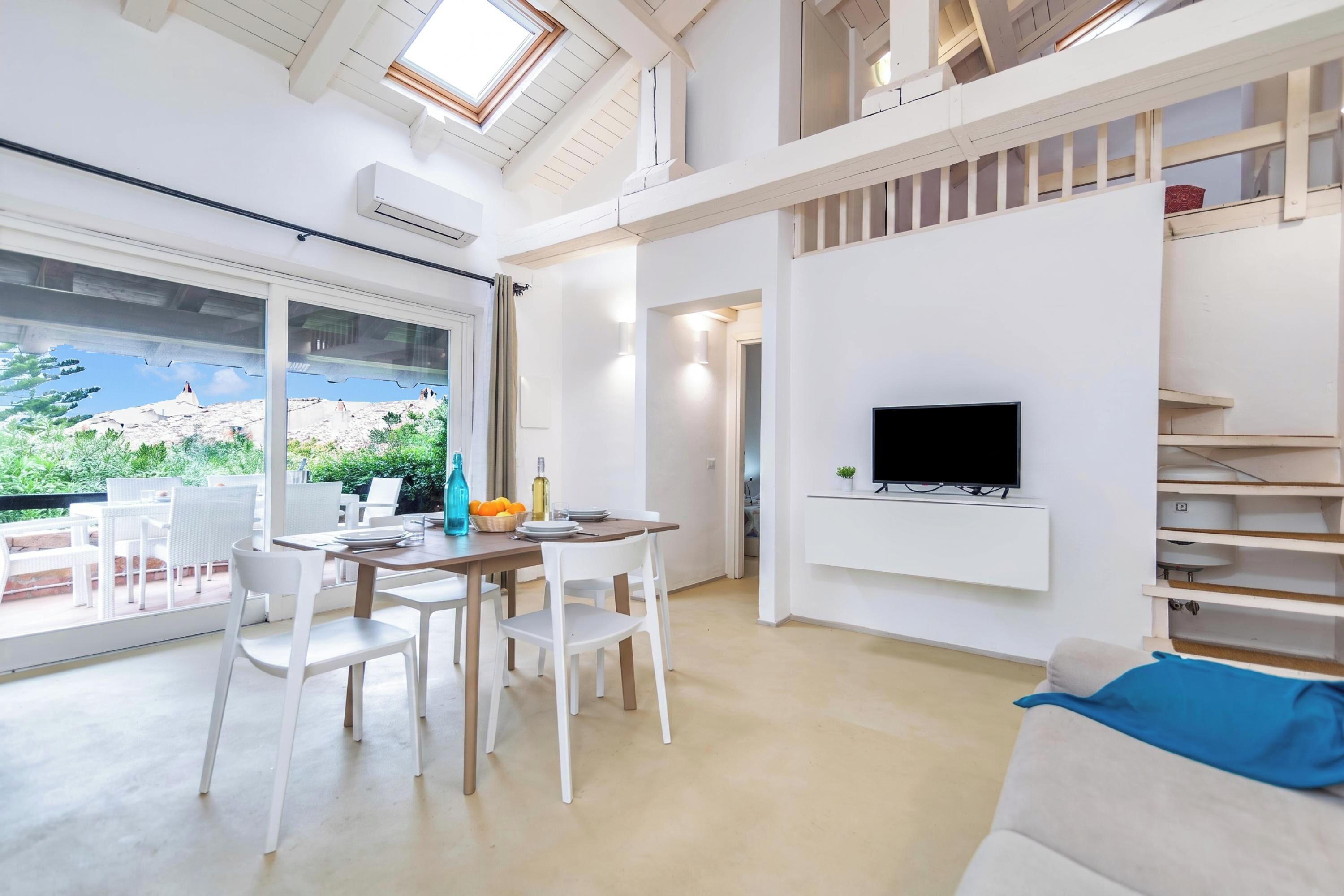Um espaço de estar moderno com área de jantar, TV fixada na parede e varanda com vista para a paisagem.