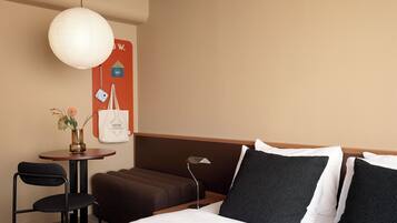 Quarto de hotel com cama, escrivaninha com cadeira, luminária pendente e placa fixada na parede.