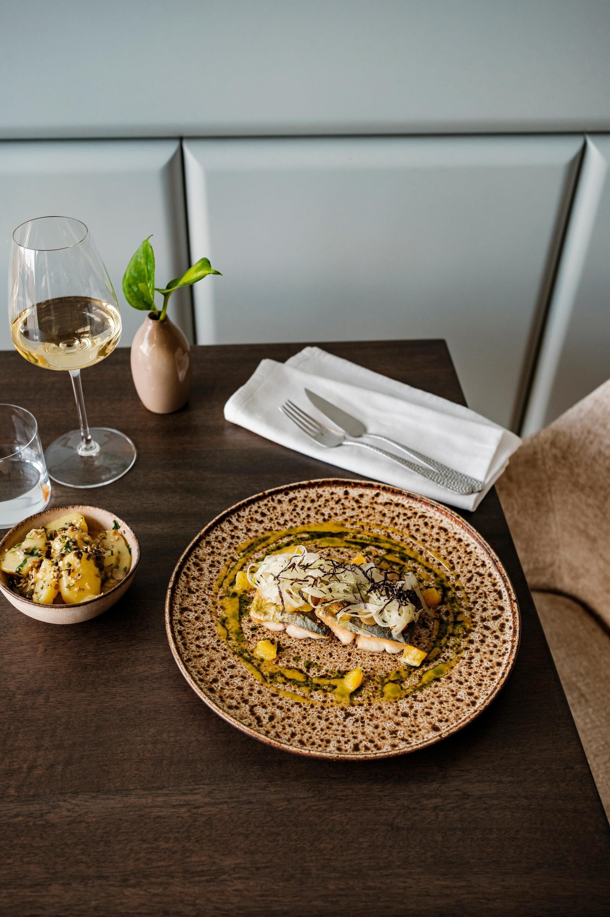 Uma mesa de jantar com um prato de comida, um copo de vinho branco e uma pequena tigela de batatas.