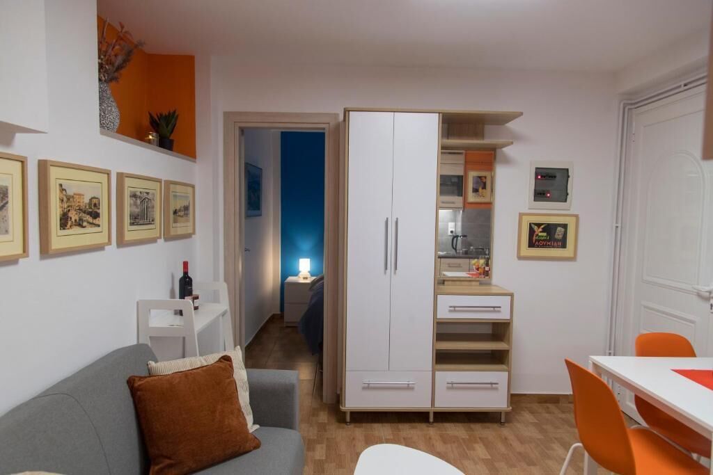 Um espaço compacto para viver, com um sofá cinzento, um guarda-roupa branco, zona de refeições com cadeiras laranja e kitchenette.