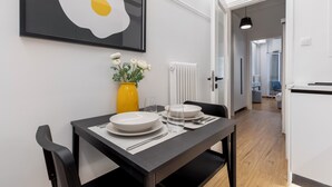 Uma área de refeições com uma mesa posta para dois, uma obra de arte emoldurada acima e um vaso amarelo com flores.