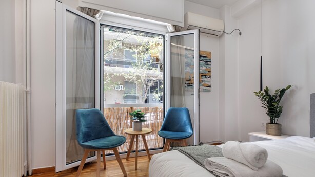 Quarto com uma cama, duas cadeiras, uma mesa pequena com uma planta e vista para o exterior por uma porta de correr.