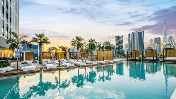 Piscina no terraço com espreguiçadeiras e palmeiras, cercada por edifícios altos e um céu claro.