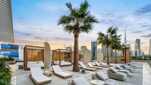 Área de lounge no terraço com espreguiçadeiras, palmeiras e o horizonte da cidade ao fundo.