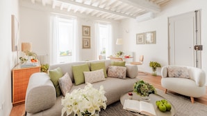 Uma sala de estar moderna com um sofá, poltronas e uma mesa de centro. Há quadros nas paredes e uma janela com cortinas.