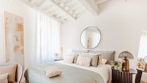 Quarto com cama, espelho redondo, mesa de cabeceira com abajur e uma cadeira.
