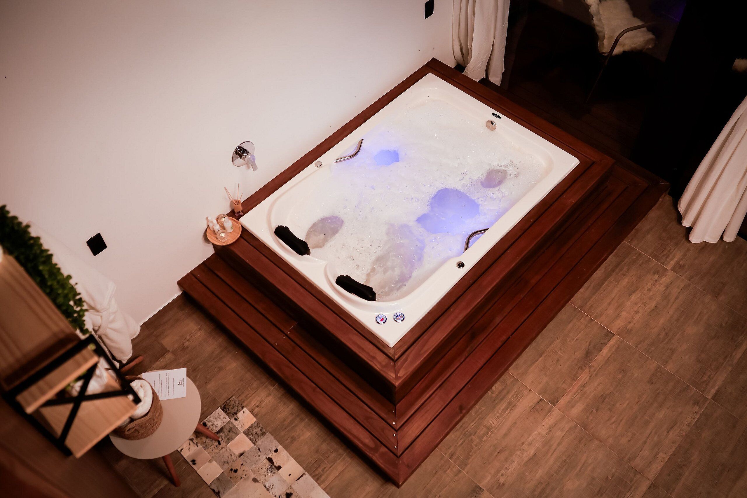 Uma banheira de hidromassagem com estrutura de madeira, uma pequena mesa com uma vela e uma planta.
