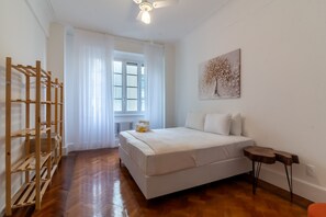 Um quarto com piso de madeira, uma cama com roupa de cama branca, uma janela com cortinas leves, uma obra de arte pendurada na parede e uma pequena mesa de madeira.