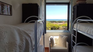 Quarto com beliches, uma cadeira pequena e janela com vista para áreas verdes e edifícios.