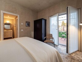 Quarto com cama, penteadeira, espelho, guarda-roupa e uma cadeira perto de uma porta de correr que dá para área externa.
