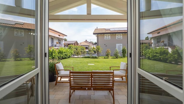 Pátio com duas bancos de madeira e porta de vidro que dá para uma área residencial com casas e gramado.