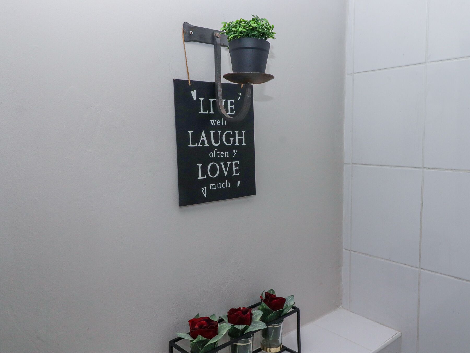 Um banheiro com uma placa preta com a inscrição "Live well, laugh often, love much" e um pequeno vaso de planta. Há três rosas vermelhas em vasos de vidro sobre um suporte de metal preto.