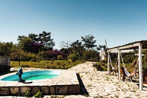 Área externa com piso de pedra, piscina, rede e pérgola.