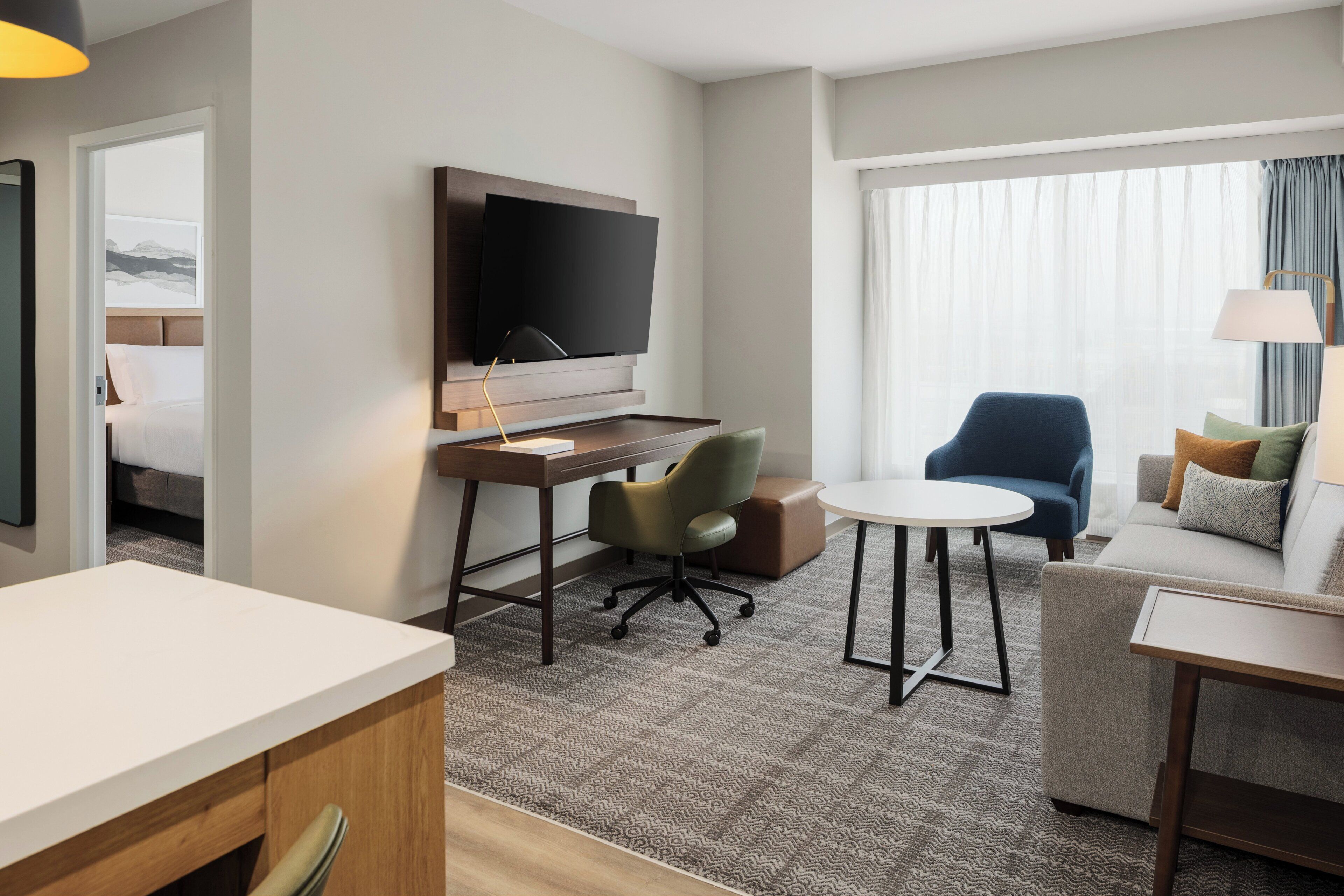 Quarto de hotel com cama, escrivaninha com televisão de tela plana, cadeira verde, poltrona azul, mesa redonda pequena e sofá com almofadas.