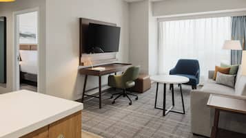 Quarto de hotel com cama, escrivaninha com televisão de tela plana, cadeira verde, poltrona azul, mesa redonda pequena e sofá com almofadas.