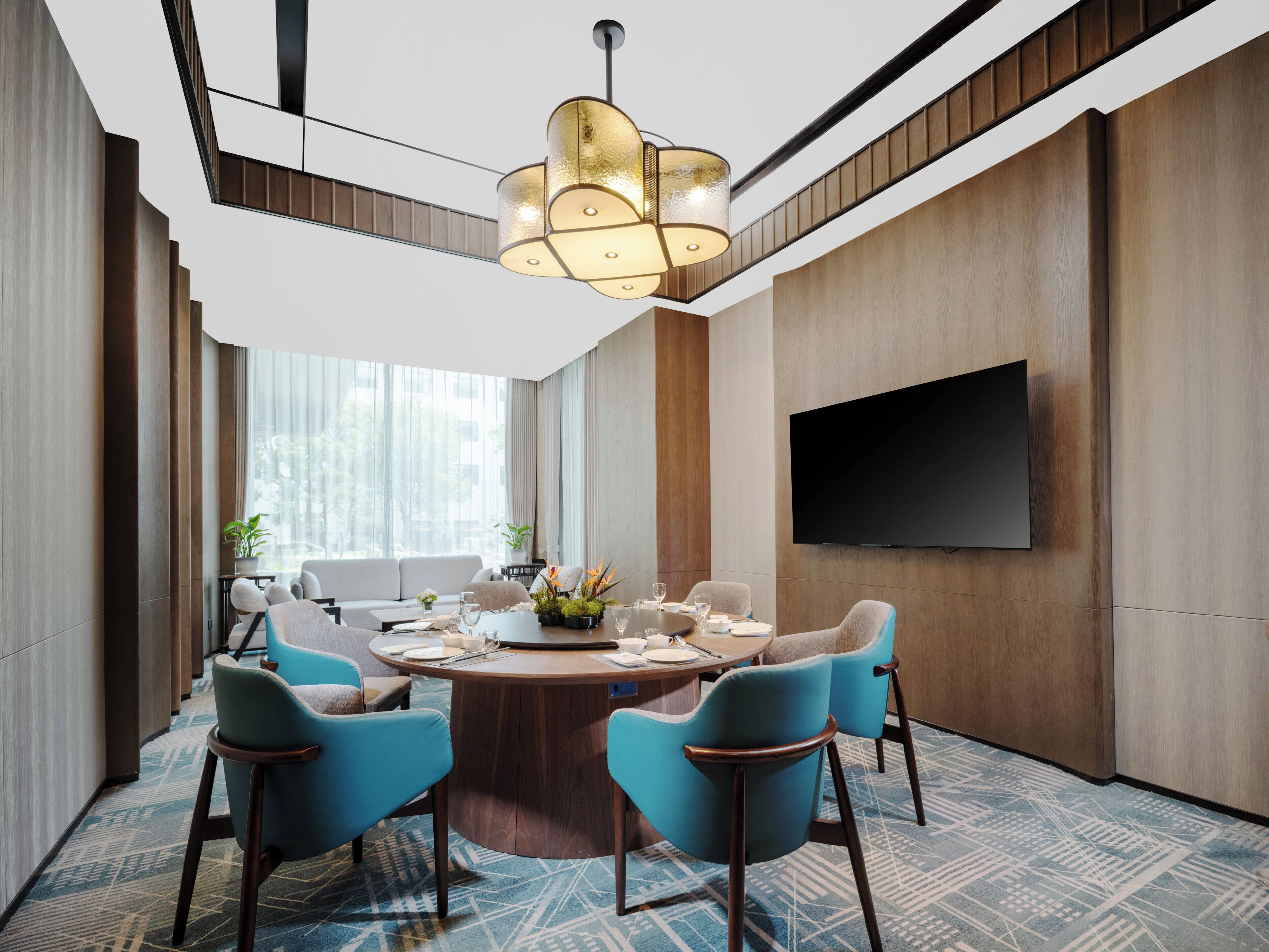 Uma sala de conferências moderna com uma mesa redonda, cadeiras azuis, um candelabro, uma TV de tela plana e uma janela grande com cortinas.