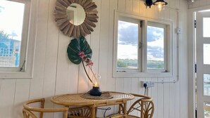 Um quarto com paredes brancas com painéis, uma mesa redonda com quatro cadeiras de vime, um espelho com moldura em forma de sol e duas luminárias fixadas na parede.