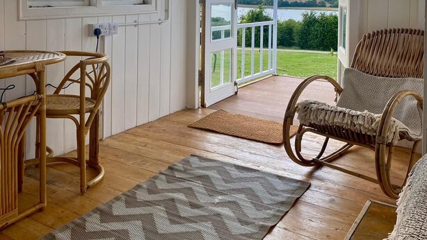 Quarto com piso de madeira, uma cadeira de vime, uma mesa e vista para o exterior.
