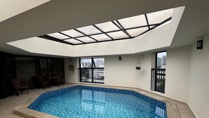 Piscina no terraço com teto de vidro, cercada por azulejos bege e grade de metal.