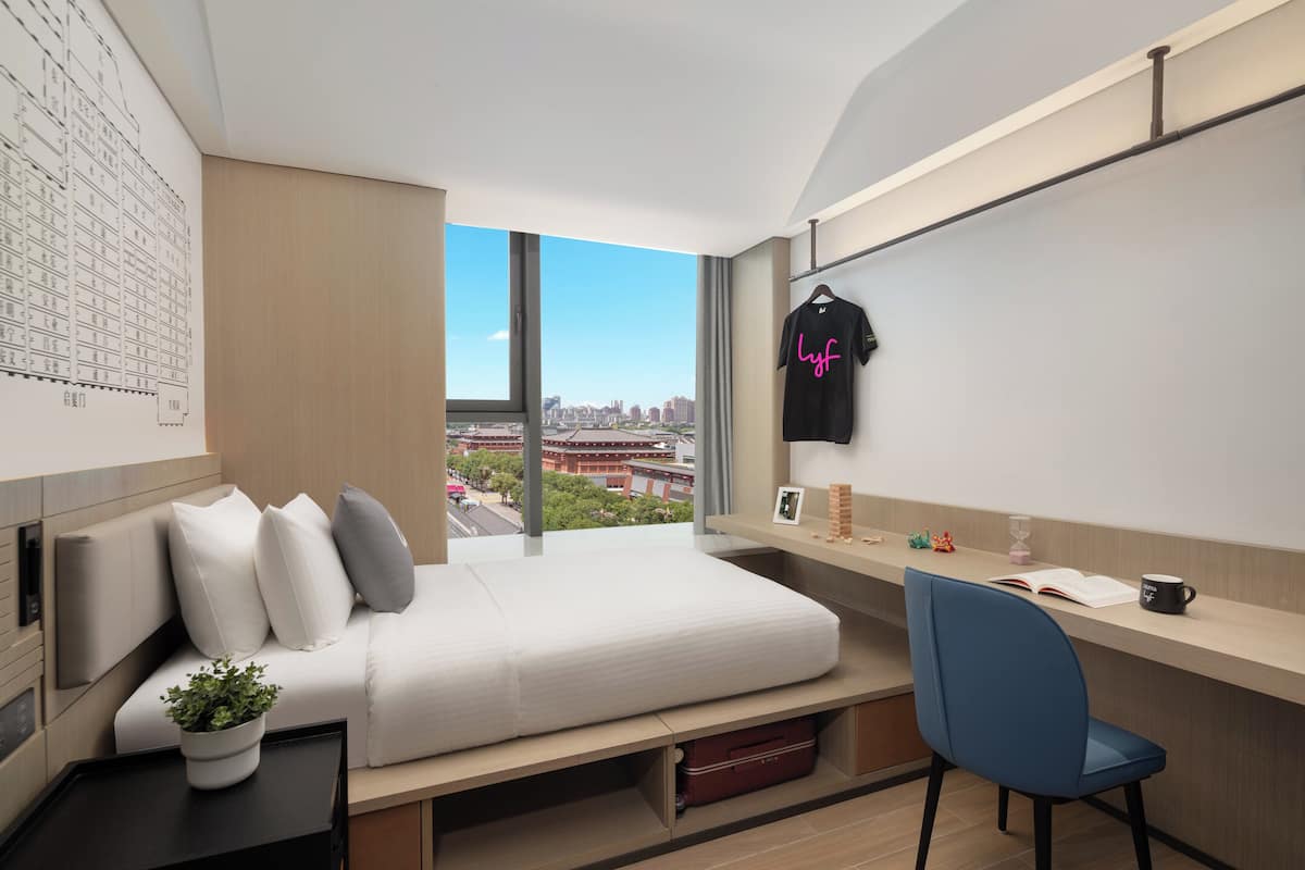 Quarto de hotel moderno com cama, escrivaninha, cadeira, planta em vaso e vista para a cidade.