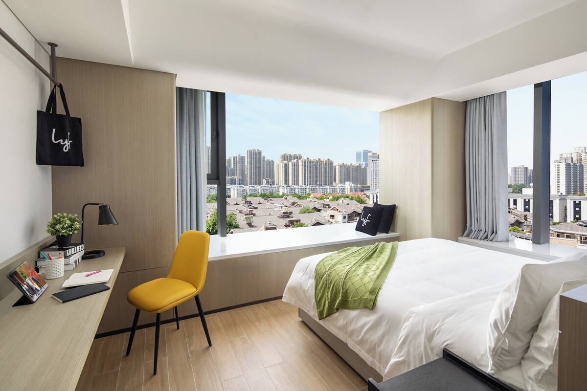 Quarto de hotel moderno com uma cama grande, uma escrivaninha com cadeira, vista do horizonte urbano através de amplas janelas e uma mala preta pendurada na parede.