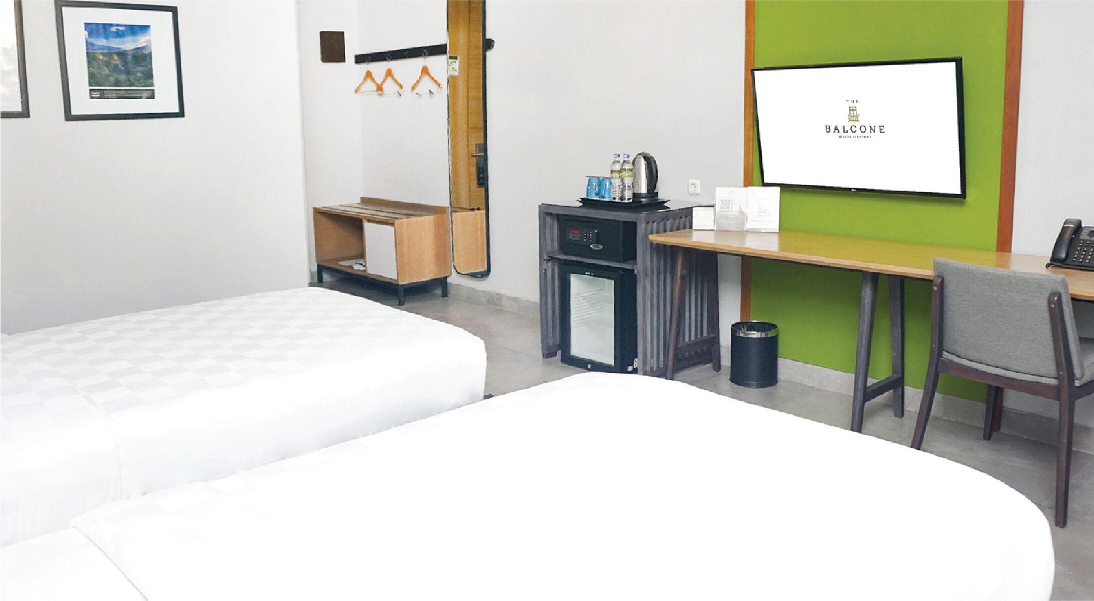 Quarto de hotel com duas camas, uma mesa com um computador, um frigorífico pequeno e uma televisão a exibir 'The Balcone'.