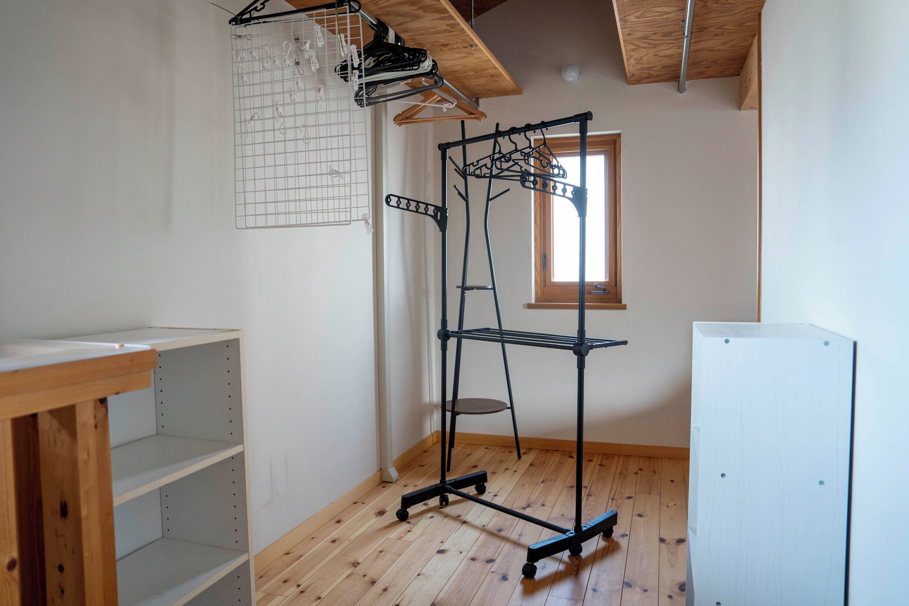 Quarto com piso de madeira, um porta-roupas de metal com roupas penduradas, uma geladeira branca e uma janela com luz natural.
