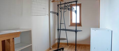 Quarto com piso de madeira, um porta-roupas de metal com roupas penduradas, uma geladeira branca e uma janela com luz natural.