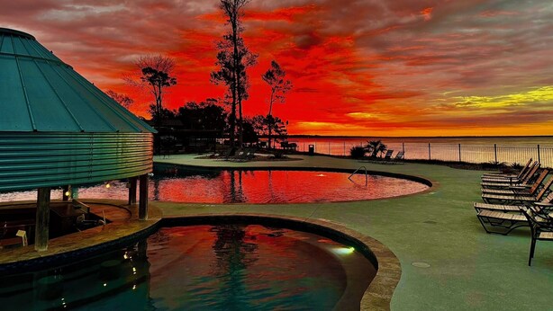 Área de piscina com vista para o pôr do sol, cadeiras de descanso e estrutura de edifício.