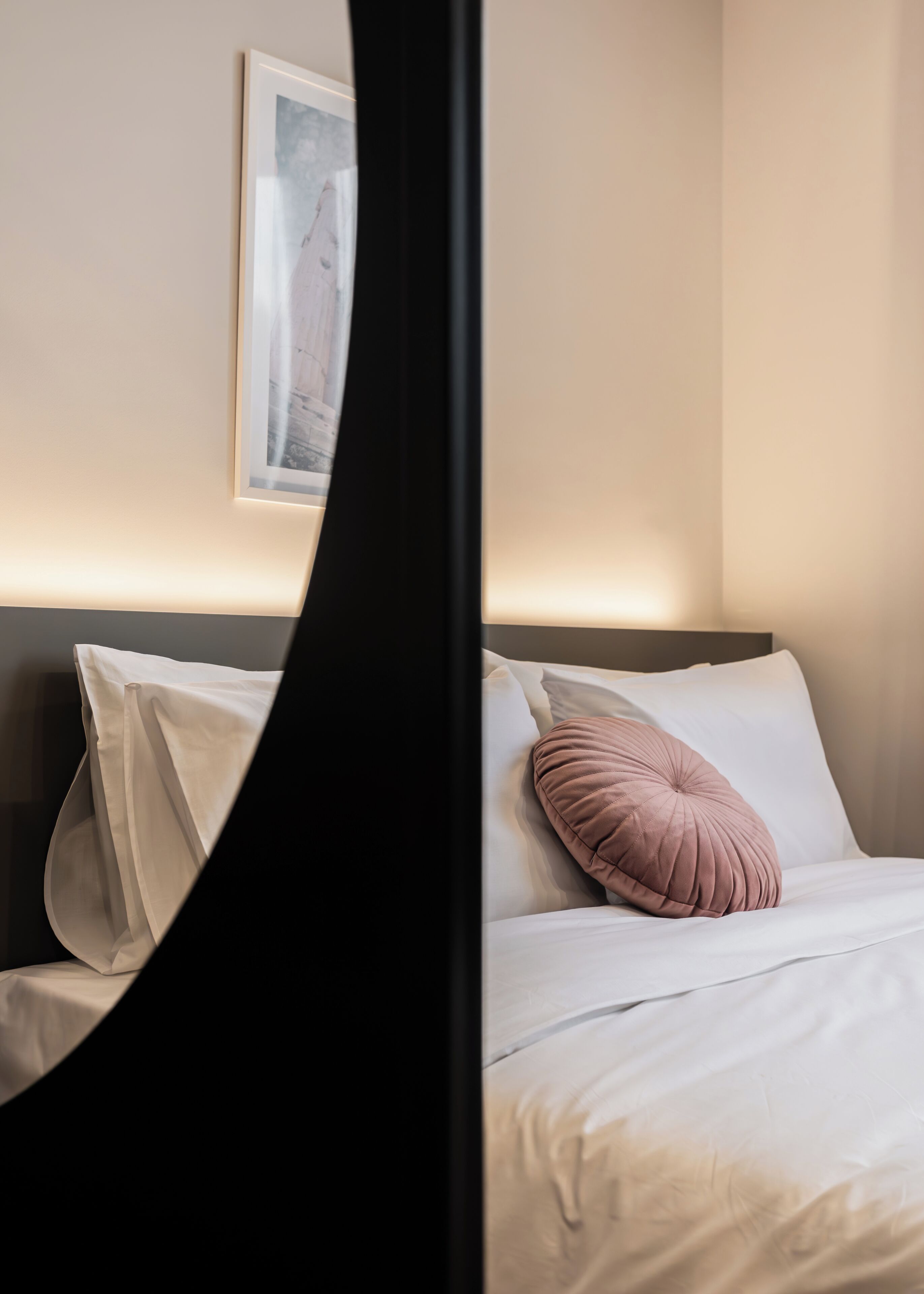 Cama bem arrumada com travesseiros brancos e cor-de-rosa, um quadro na parede e um objeto escuro que cobre parcialmente a vista.