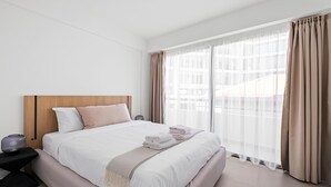 Quarto de hotel moderno com uma cama grande, uma mesinha de cabeceira e vista para a cidade através de cortinas translúcidas.