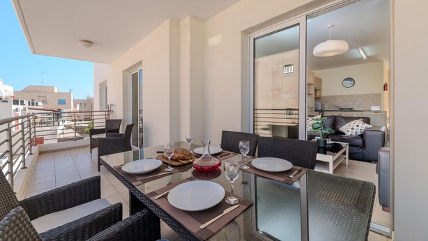 Uma sala de jantar com mesa posta para quatro pessoas, um sofá e cozinha ao fundo.