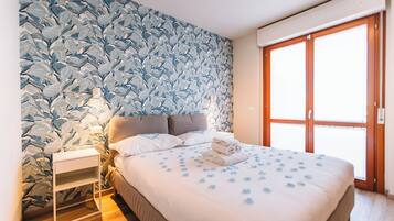 Quarto com papel de parede floral, uma cama com toalhas dobradas, uma mesa de cabeceira e uma janela grande.