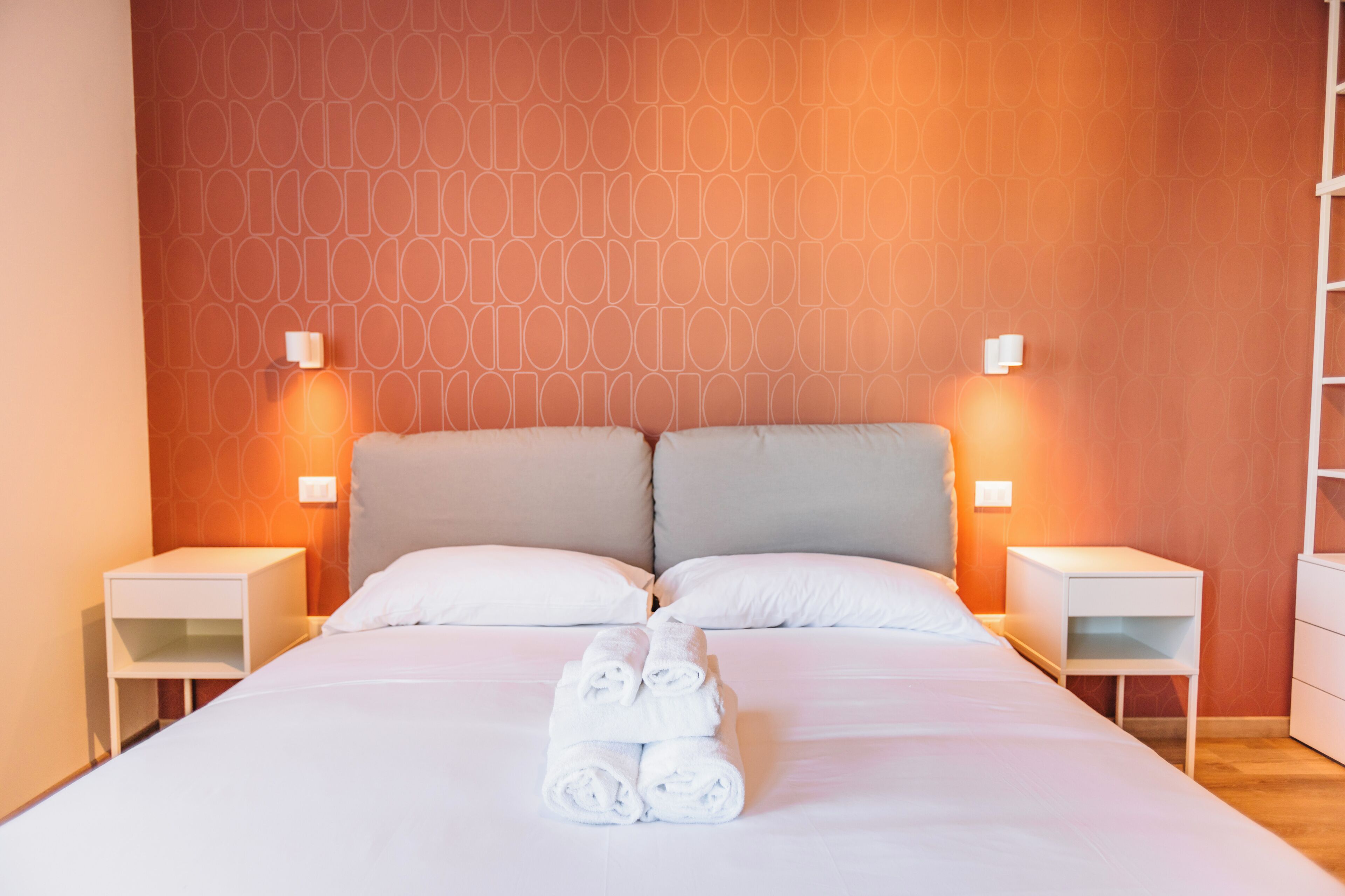 Cama bem arrumada com roupa de cama e toalhas brancas, duas mesas de cabeceira e parede com padrão.