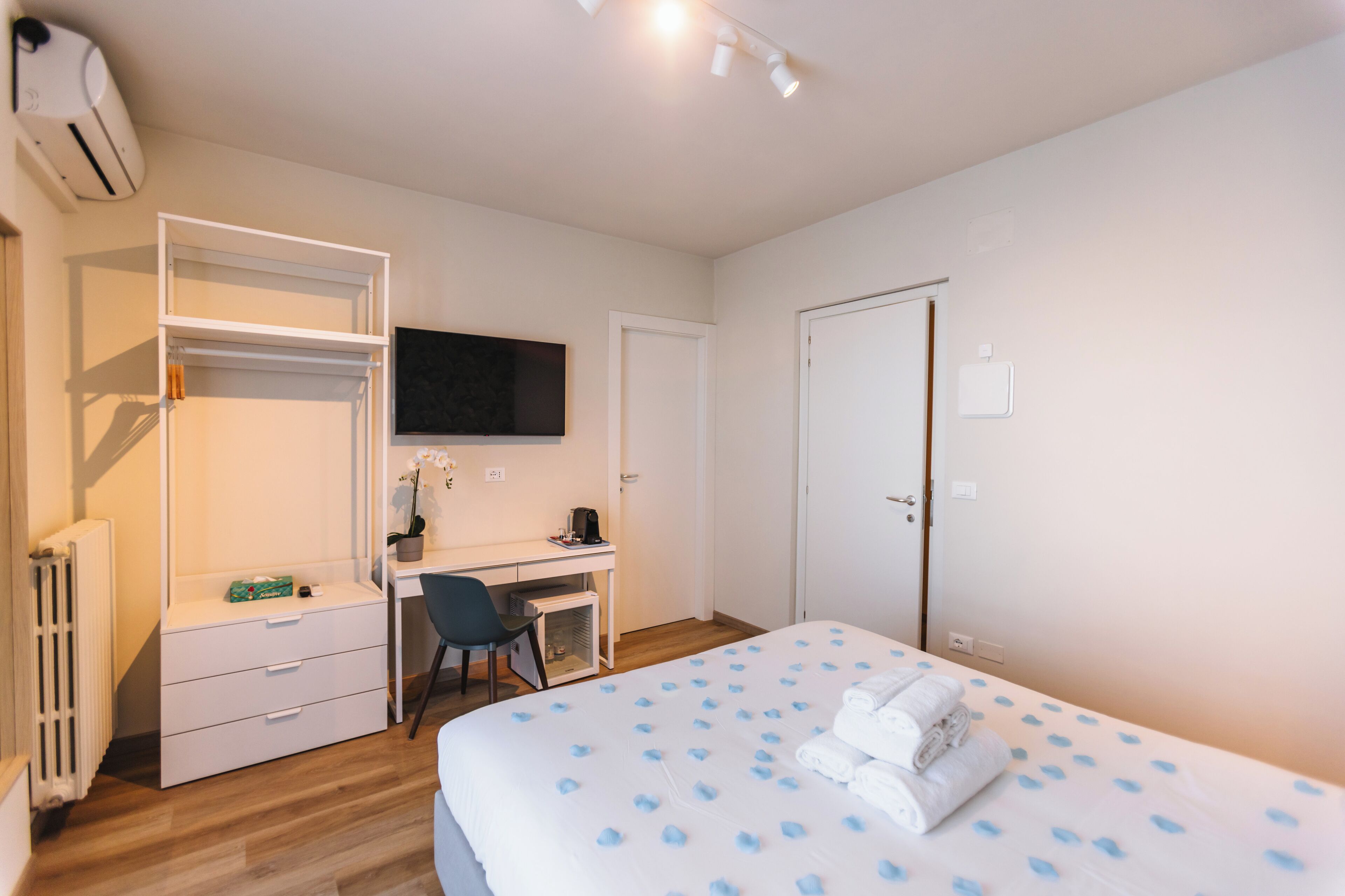 Quarto de hotel com cama, escrivaninha, cadeira, guarda-roupa e televisão na parede.